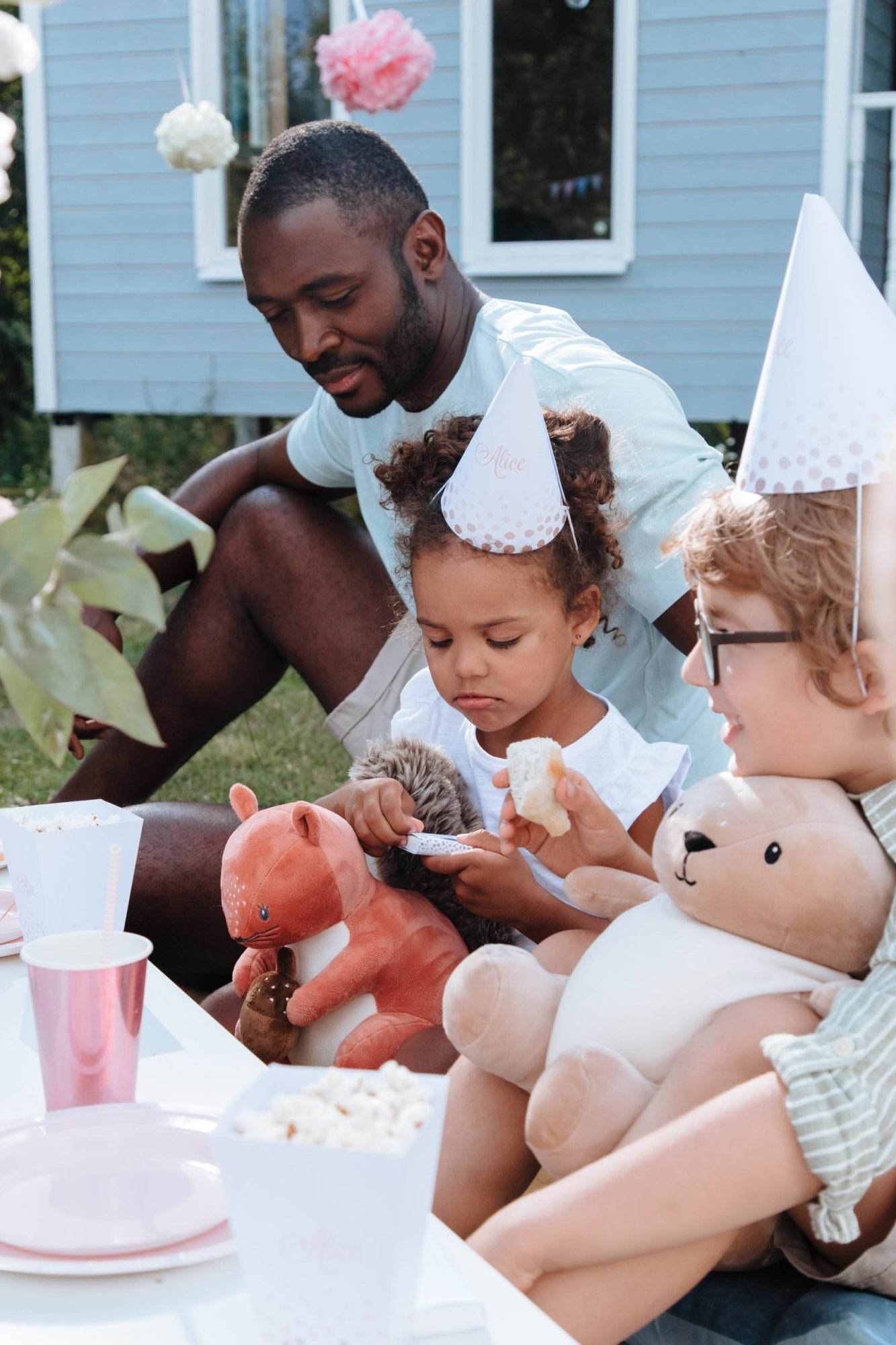 A Man And Two Children In Party Hats Sit At A Table Outdoors, Enjoying ...